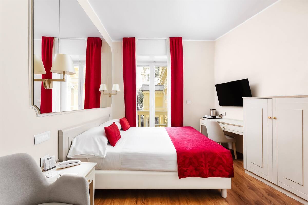 Quarto de hotel com cama, escrivaninha, cadeira e televisão.