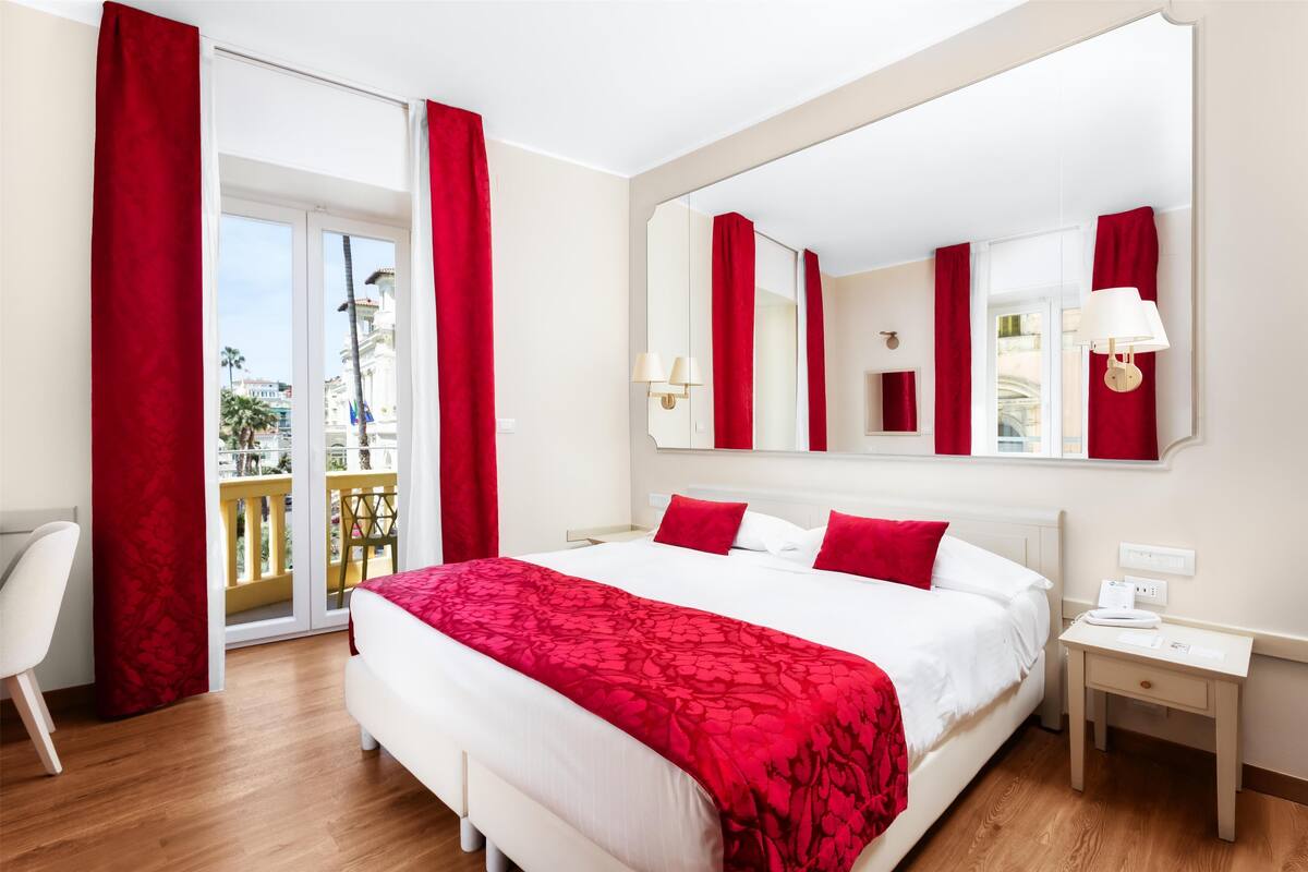 Quarto de hotel com uma cama grande, cortinas vermelhas, varanda com guarda-corpos amarelos e uma mesa de cabeceira branca.