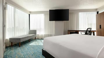 Quarto de hotel com uma cama grande, televisão de tela plana na parede, uma escrivaninha com cadeira e um banco perto da janela.
