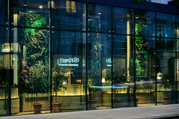 Fachada moderna de um edifício com grandes janelas de vidro refletindo áreas verdes e um letreiro para 'Pampero Italian Bar & Restaurant'.