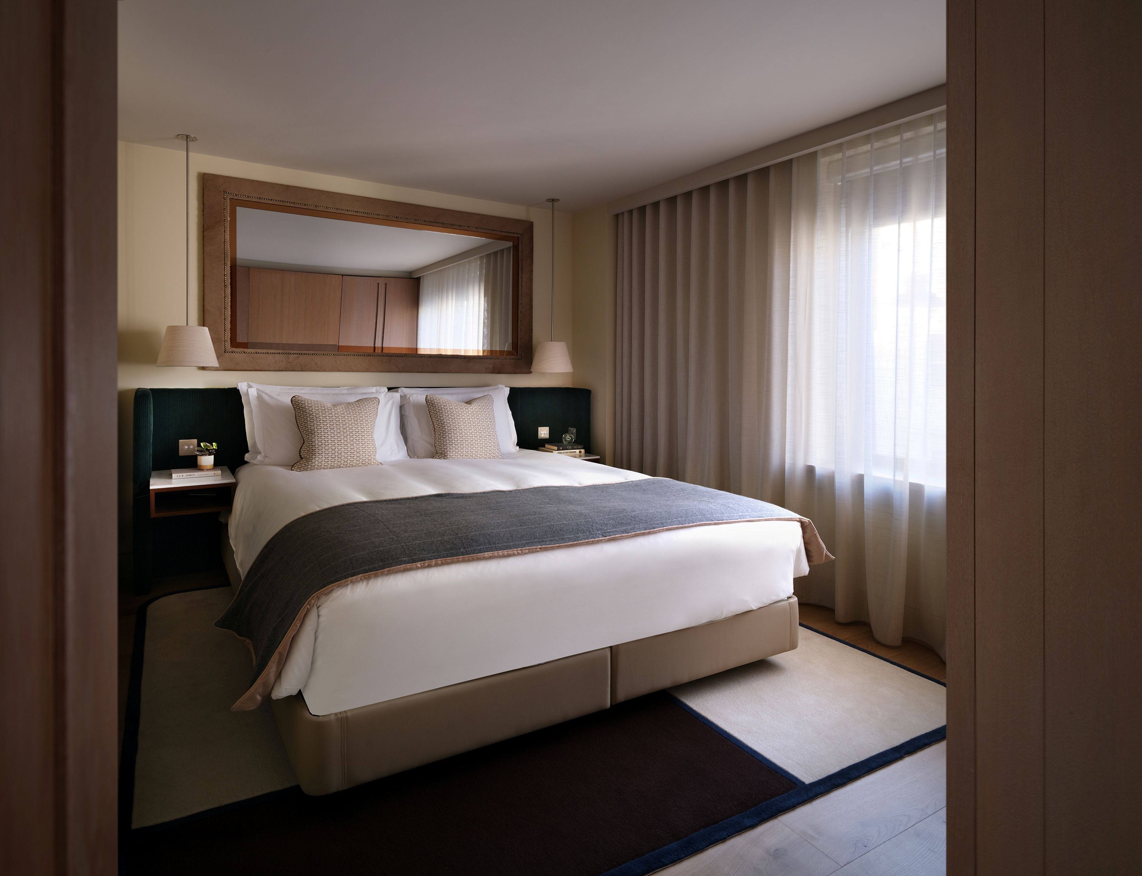 Quarto de hotel com uma cama grande, mesa de cabeceira com luminária, espelho sobre a cama e janela com cortinas.