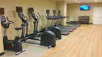 Uma academia com várias esteiras, bicicletas ergométricas e uma televisão na parede.