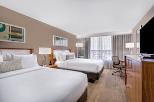 Quarto de hotel com duas camas, uma escrivaninha, uma cadeira, uma televisão e obras de arte nas paredes.
