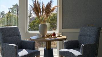 Quarto com duas poltronas azuis, uma mesa redonda com um vaso de plantas secas e vista para árvores através de grandes janelas.