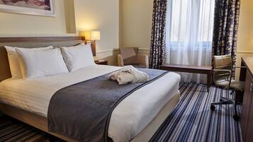 Quarto de hotel com uma cama grande, uma escrivaninha, uma cadeira e uma janela com cortinas.