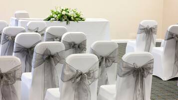 Fileiras de cadeiras brancas com fitas cinzas em laços, dispostas em um salão de festas com um arranjo floral sobre uma mesa.