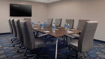Uma sala de conferências com uma mesa grande, oito cadeiras, uma TV de tela plana e garrafas de água sobre a mesa.