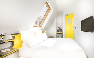 Quarto de hotel com cama, secretária com abajur, televisão, porta amarela e janela com moldura de madeira.
