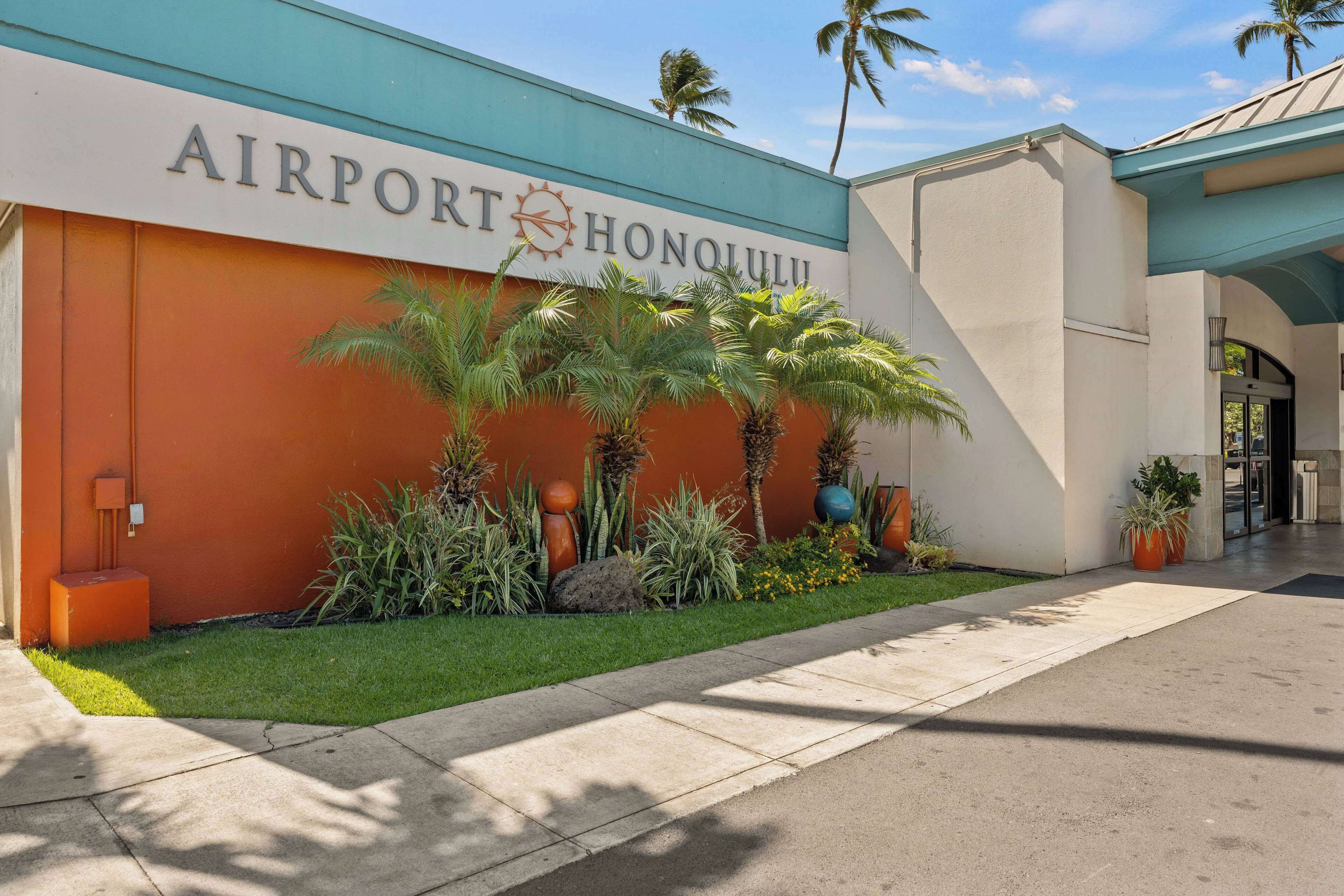 Exterior do edifício do Aeroporto Honolulu, com uma placa, palmeiras e um gramado bem cuidado.