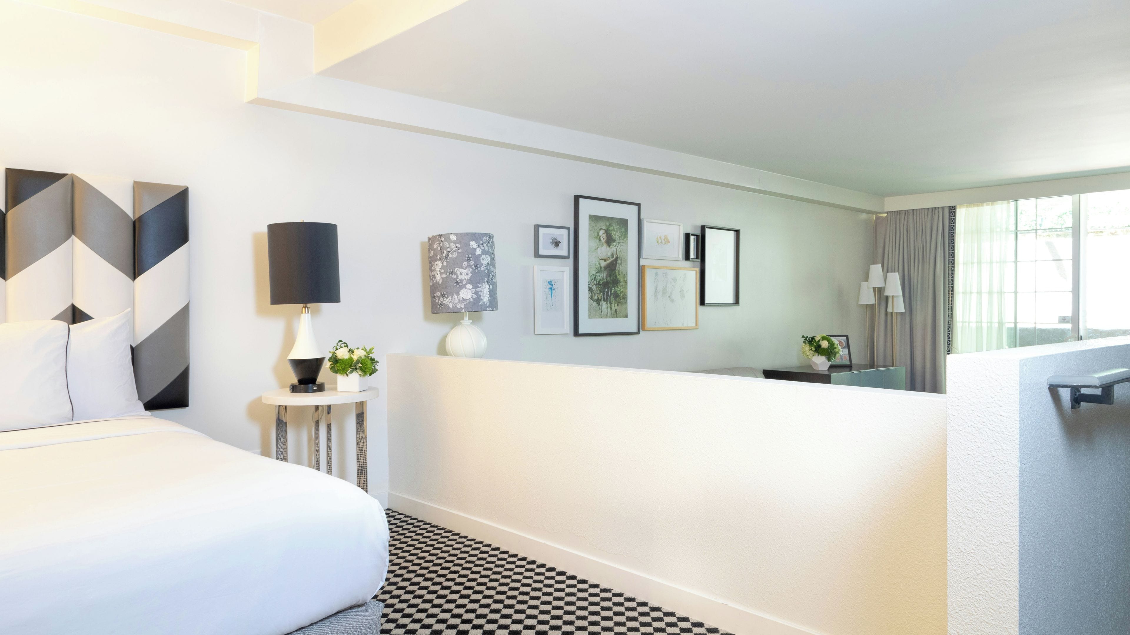 Quarto de hotel com uma cama grande, uma mesinha com abajur, parede com quadros e uma janela com cortinas.