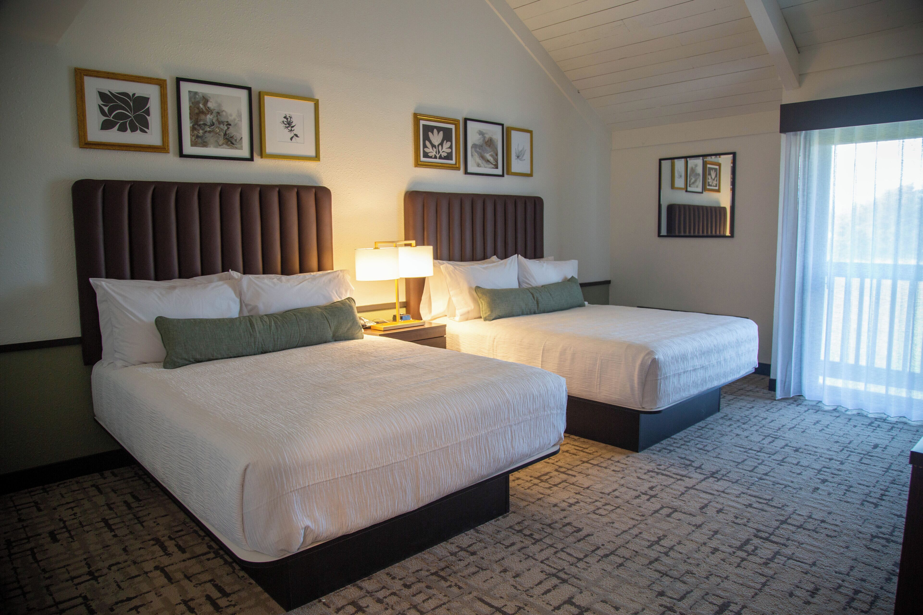 Quarto de hotel com duas camas, mesa de cabeceira com abajur e quadros nas paredes.