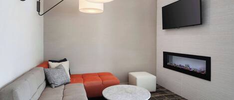 Uma sala de estar moderna com um sofá cinza, assentos laranja como detalhe, uma mesa de centro redonda, uma TV de tela plana na parede e um tapete estampado.
