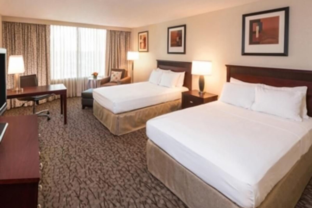Quarto de hotel com duas camas, uma escrivaninha, uma cadeira e um abajur.