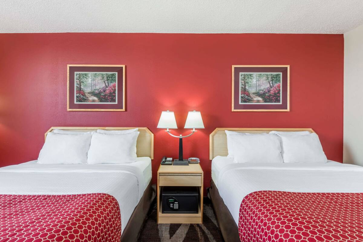 Quarto de hotel com duas camas, uma mesa de cabeceira com abajur e duas fotos emolduradas na parede.
