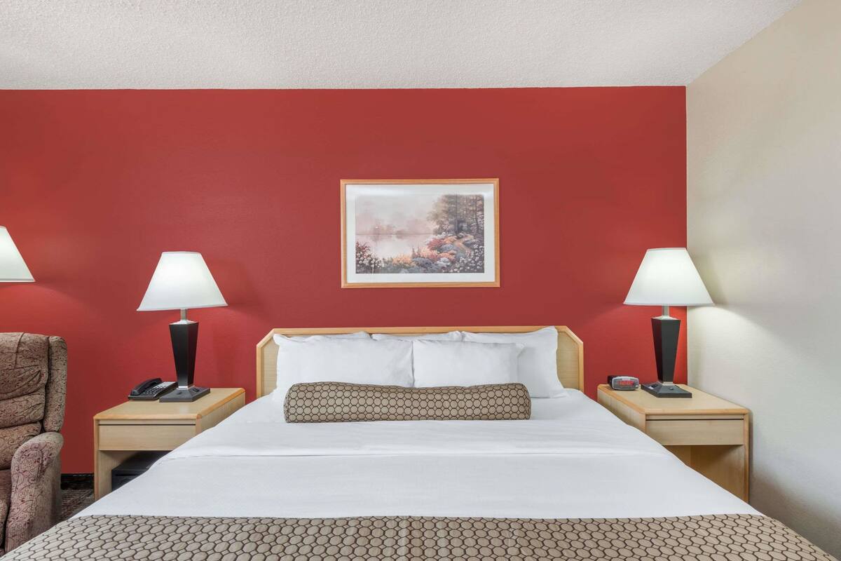 Quarto de hotel com uma cama, duas mesas de cabeceira com abajures, uma cadeira e um quadro na parede.