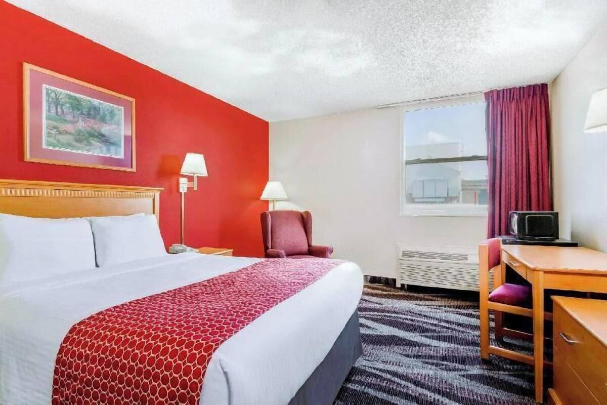 Quarto de hotel com cama, escrivaninha, cadeira, televisão, abajur e um quadro na parede.