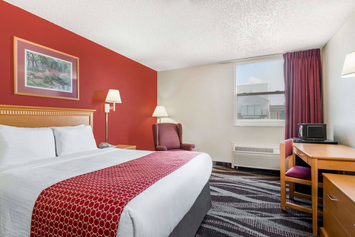 Quarto de hotel com uma cama grande, escrivaninha, cadeira, televisão e janela com cortinas.