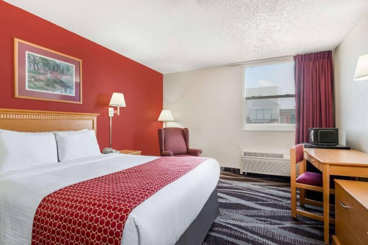 Quarto de hotel com cama, escrivaninha, cadeira, televisão, janela com cortinas e um quadro na parede.