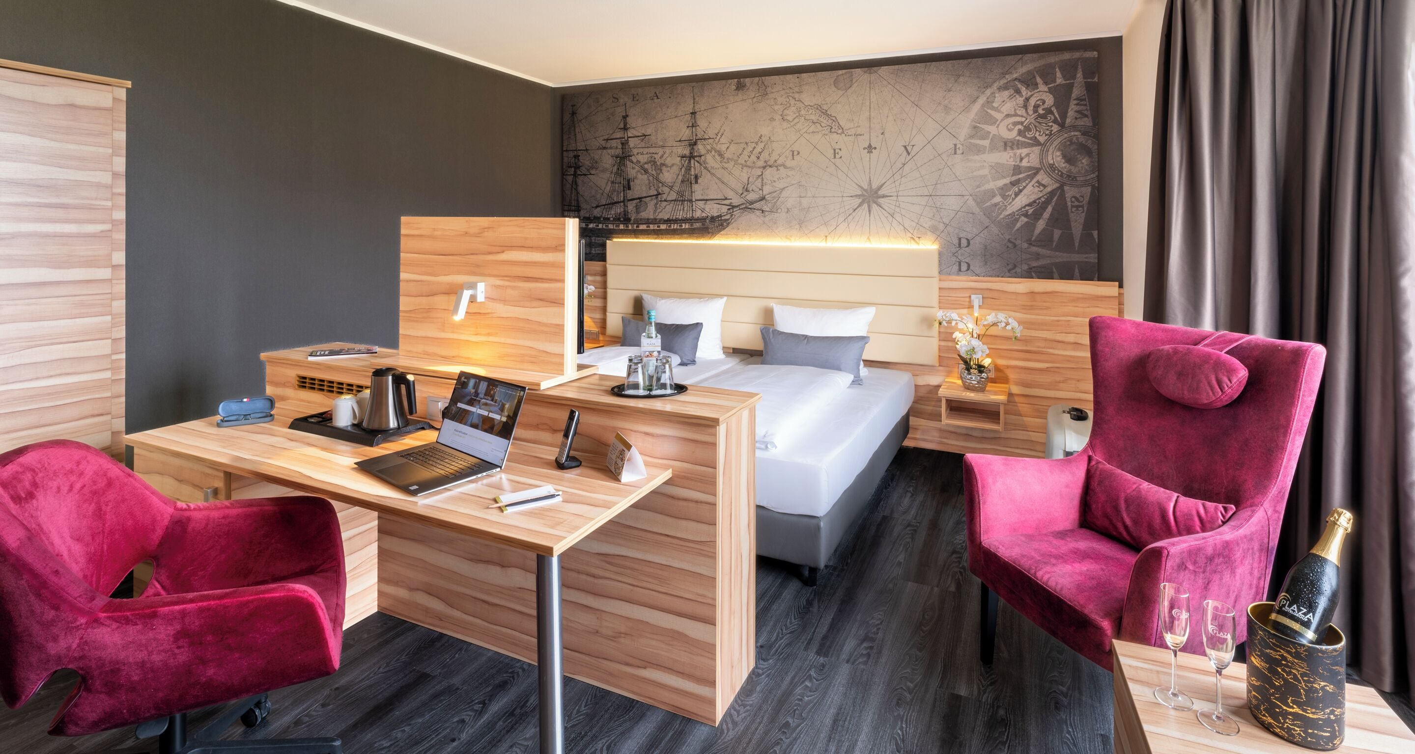 Quarto de hotel moderno com escrivaninha de madeira, cama com roupa de cama branca, poltrona de veludo vermelho e um mapa grande na parede.