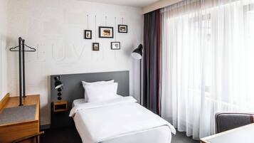 Quarto de hotel com cama, escrivaninha, cadeira, mesa de cabeceira, abajur e janela com cortinas.