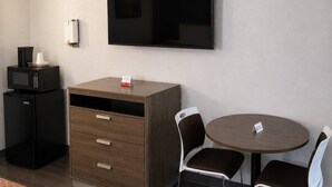 Quarto de hotel com televisão de tela plana fixada na parede, escrivaninha de madeira com gavetas, pequeno frigorífico preto, micro-ondas, duas cadeiras marrons ao redor de uma mesa redonda e luminária branca presa à parede.