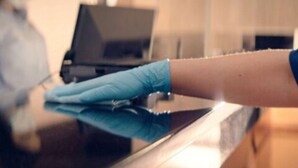 Uma pessoa usando luvas azuis limpando uma superfície refletora, provavelmente uma mesa ou bancada.