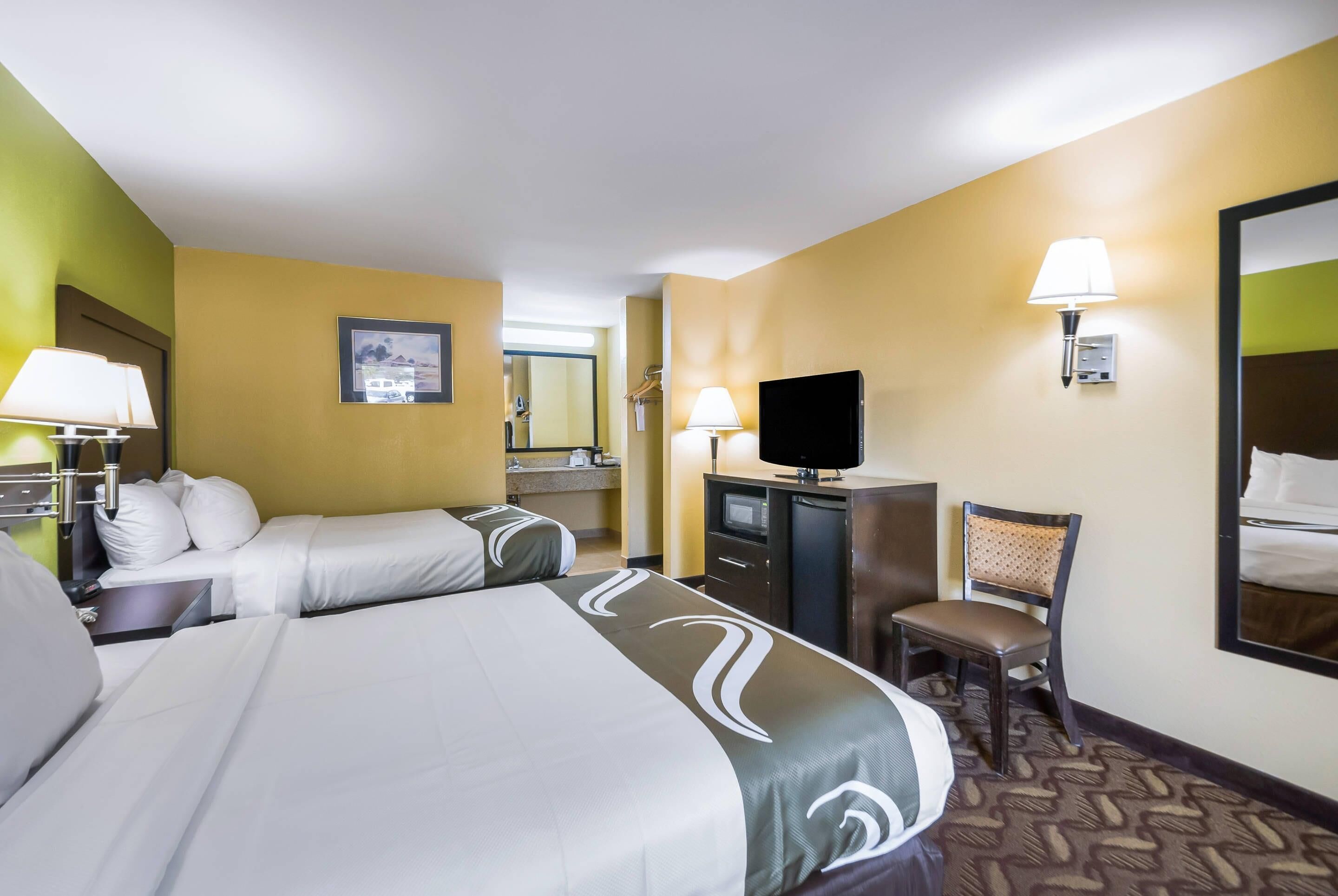 Quarto de hotel com duas camas, uma escrivaninha, uma cadeira, uma televisão e um espelho.