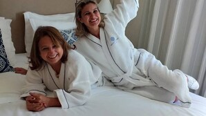 Duas pessoas vestidas de branco sobre uma cama com roupa de cama branca, uma cabeceira de madeira e um espelho acima da cama.