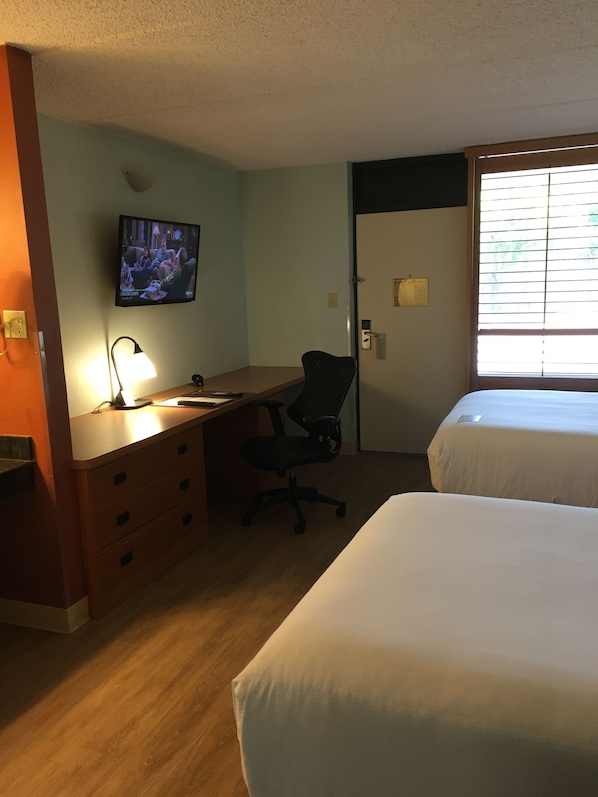 Quarto de hotel com escrivaninha de madeira, uma cadeira, televisão fixada na parede, cama e janela com persianas.