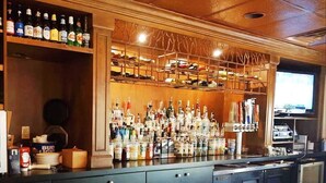 Uma área de bar com prateleiras de garrafas de bebidas, uma televisão e banquetas.