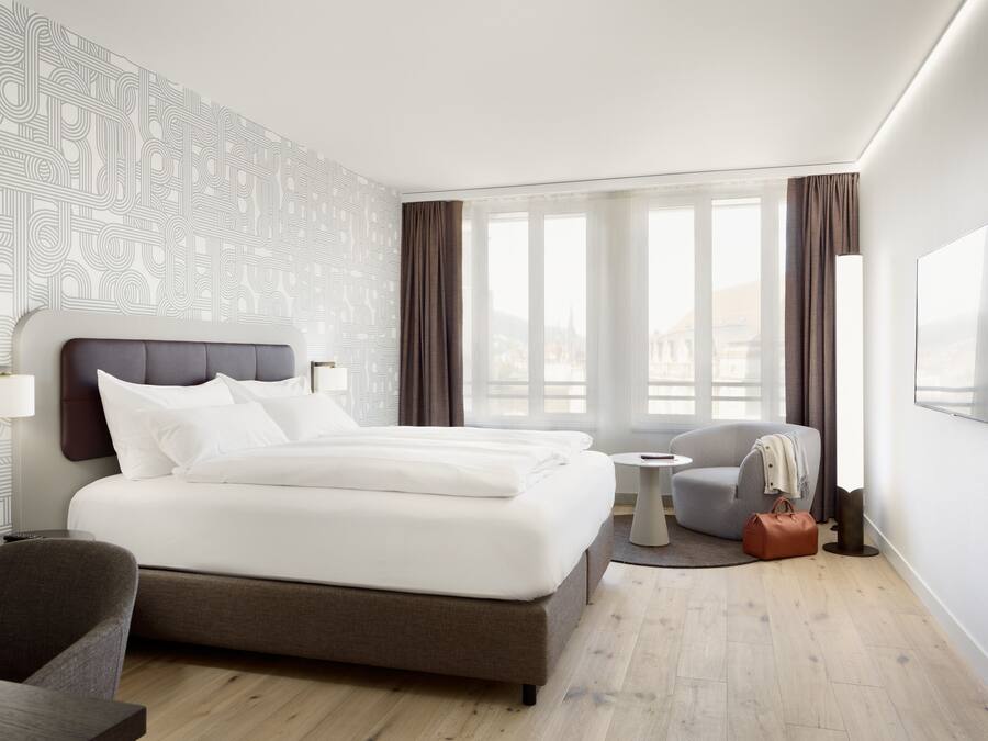 Quarto de hotel moderno com uma cama grande, área de estar e vista para a cidade.