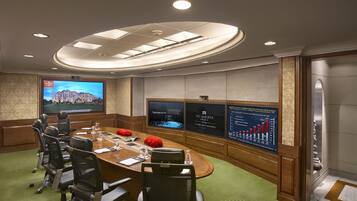 Uma sala de conferências com uma grande mesa oval, várias cadeiras, uma tela de televisão e dois grandes ecrãs digitais.