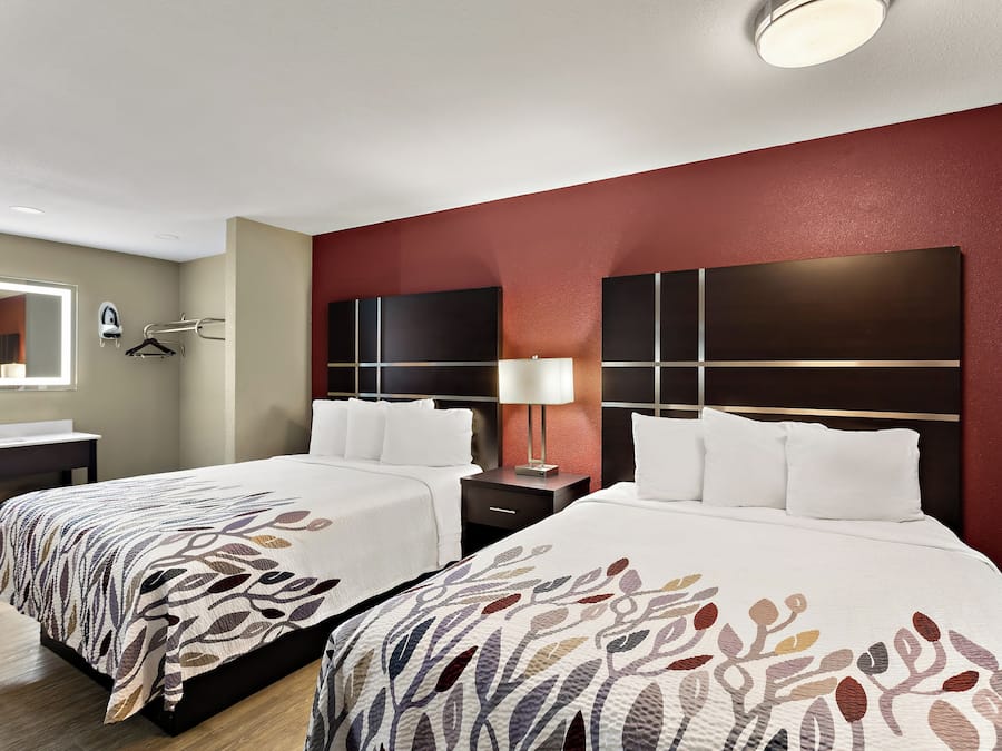 Quarto de hotel com duas camas, um espelho grande, mesa de cabeceira com abajur e uma parede de destaque vermelha.