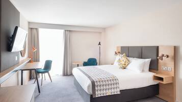 Quarto de hotel com cama, escrivaninha, cadeira, televisão e janela com cortinas.