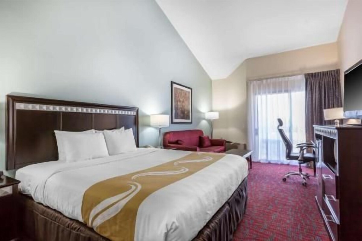 Quarto de hotel com uma cama grande, escrivaninha, cadeira, televisão e janela com cortinas.