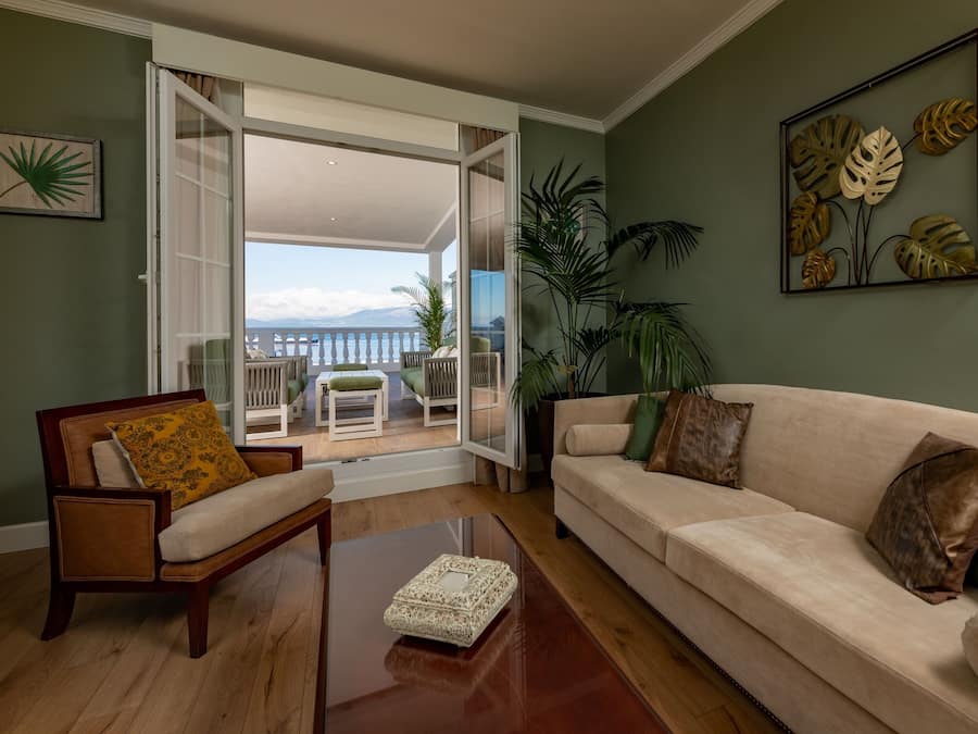 Sala de estar com sofá bege, uma cadeira de madeira e mesa de centro de vidro. Há uma varanda com vista para o mar e montanhas.