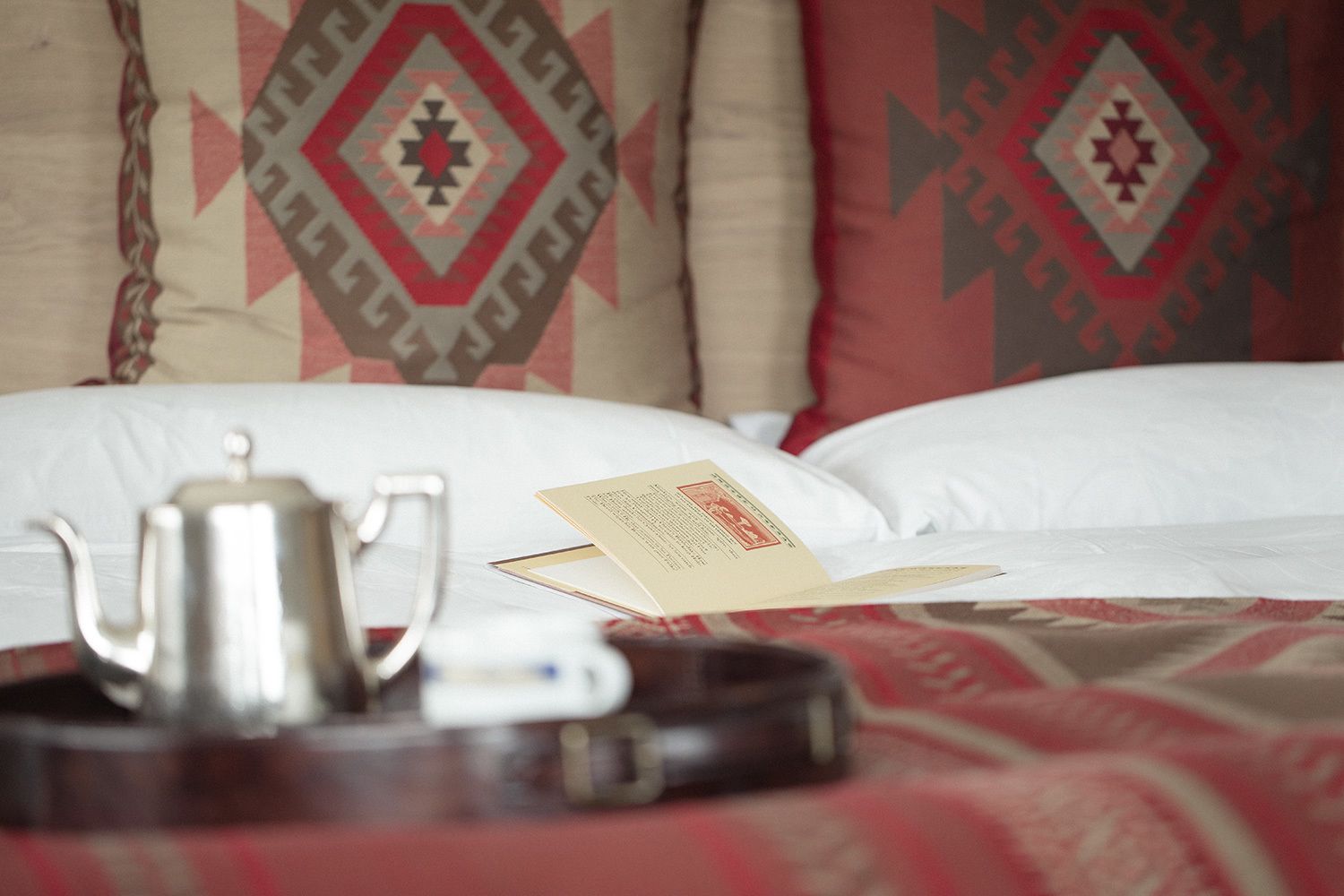 Uma cama com almofadas estampadas, um tabuleiro com bule de chá e um cardápio sobre a cama.