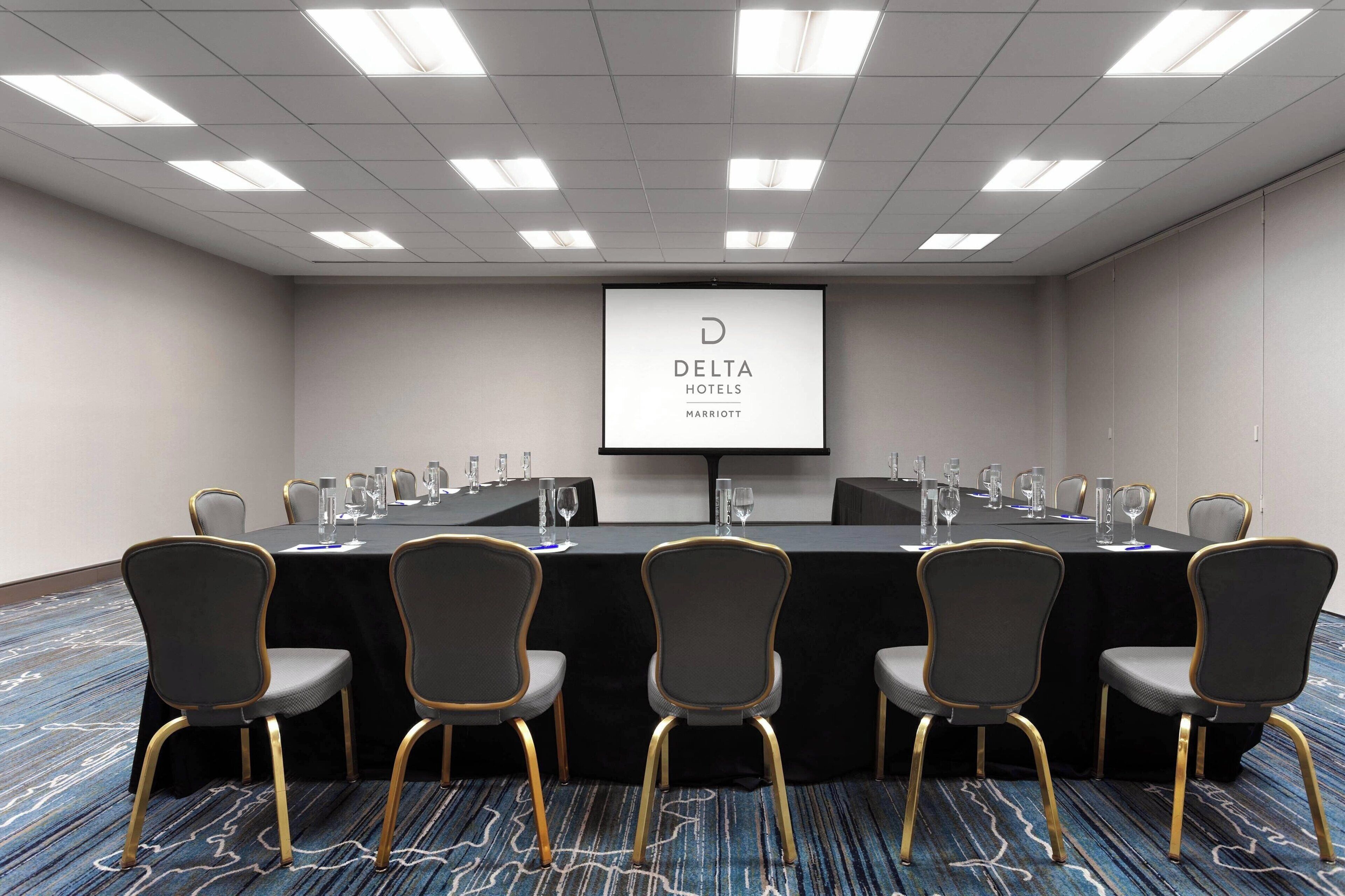 Uma sala de conferência com uma mesa comprida, seis cadeiras e uma tela de projetor exibindo "DELTA HOTELS MARRIOTT".