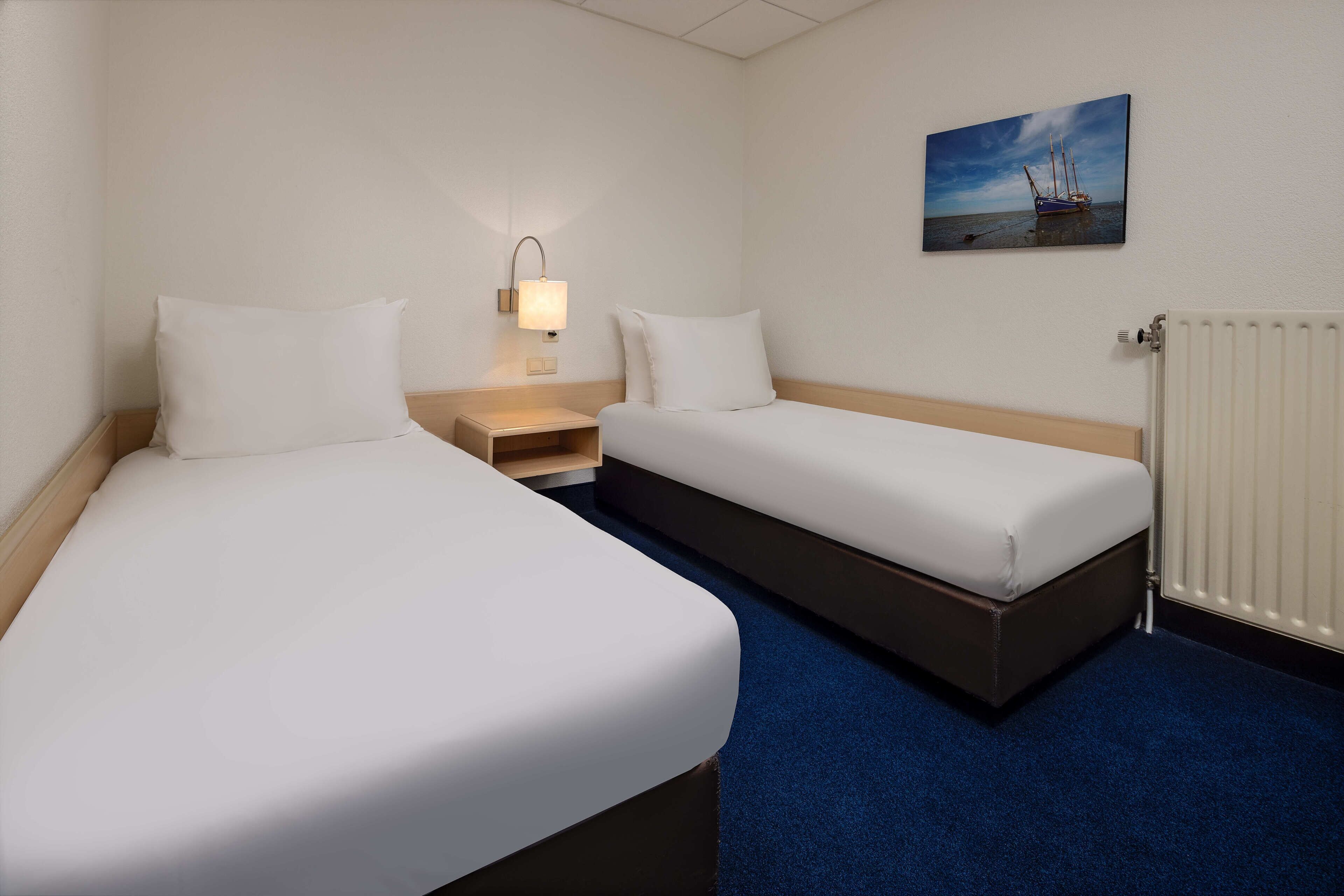 Quarto de hotel com duas camas de solteiro, uma mesinha de cabeceira, uma luminária na parede e um quadro de um veleiro.