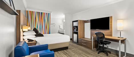 Quarto de hotel com cama, escrivaninha, cadeira, televisão e parede listrada e colorida.