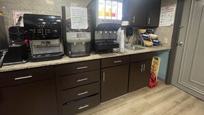 Cozinha com máquina de café, dispensador de bebidas e pia. Há armários e uma porta visíveis.