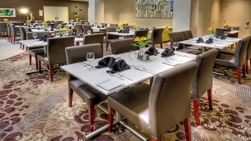 Espaço de refeições de hotel com várias mesas preparadas para hóspedes, piso com carpete e decoração moderna.
