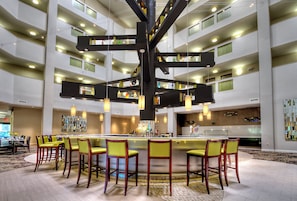 Um hall de hotel espaçoso, com bar central, diversos espaços de estar e pé direito alto com iluminação natural.