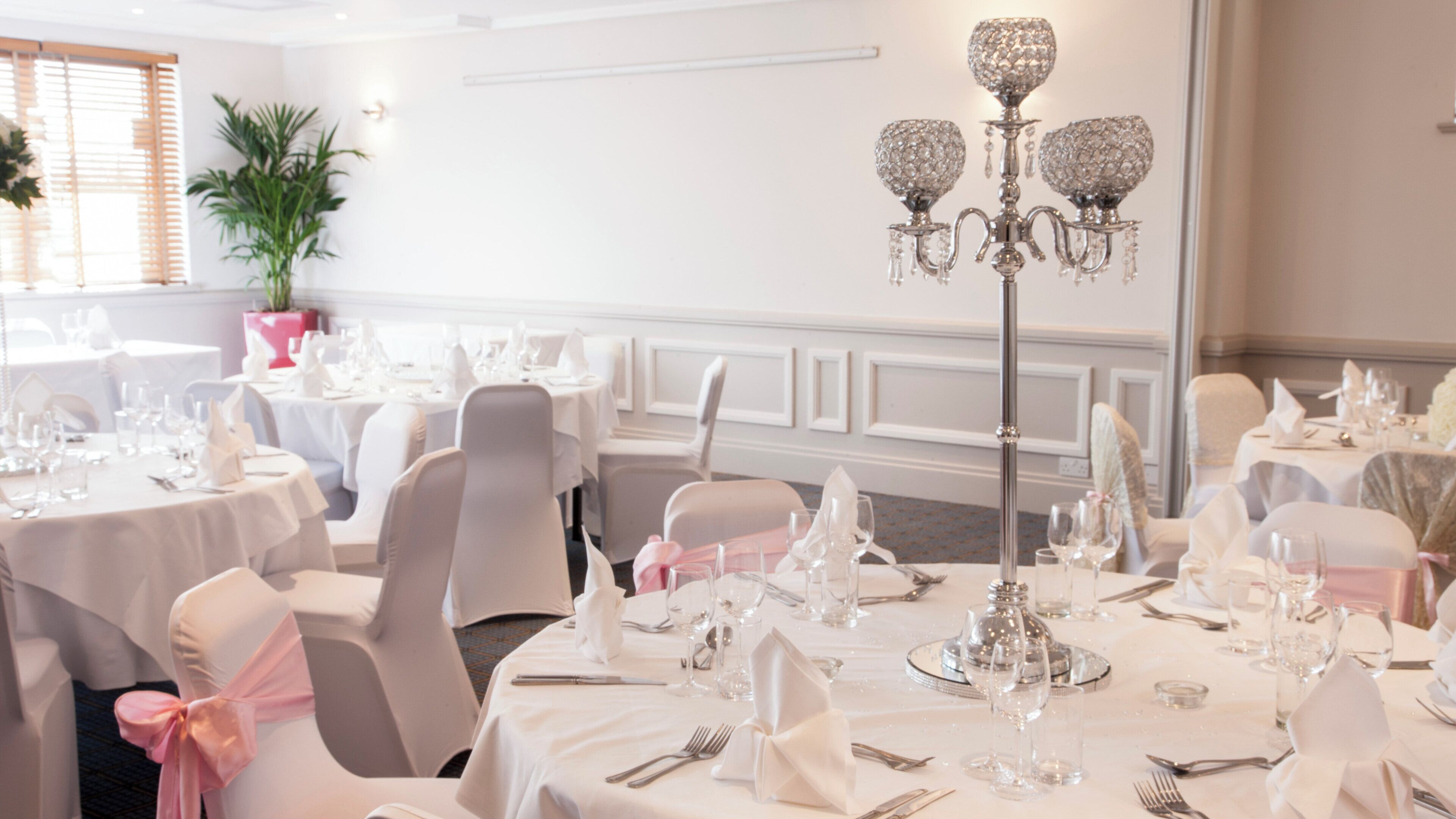 Salão de banquetes com mesas redondas montadas para refeições, cadeiras brancas com fitas cor-de-rosa e um lustre central.