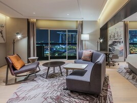 Quarto de hotel moderno com uma janela grande que oferece vista da cidade, um sofá, uma poltrona, uma mesa pequena e um abajur.