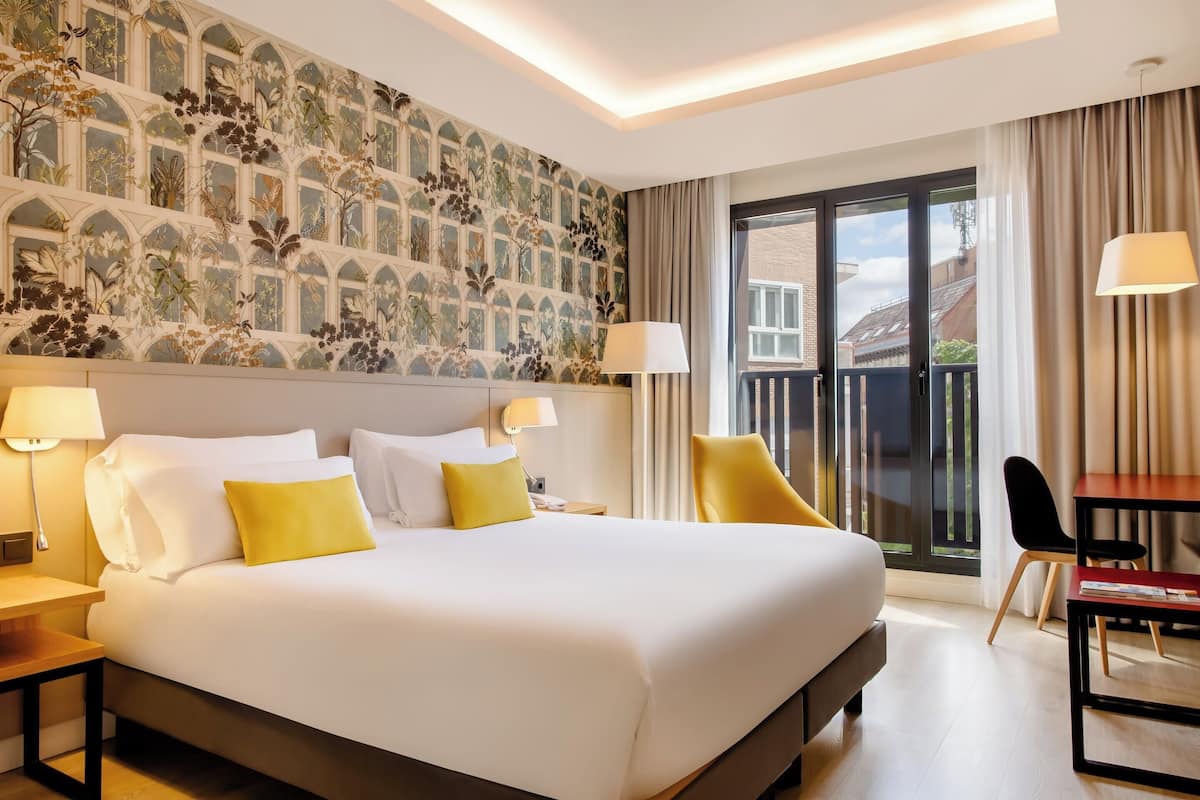 Quarto de hotel com uma cama grande, duas mesas de cabeceira, uma escrivaninha com cadeira e varanda com cortinas.