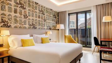 Quarto de hotel com uma cama grande, duas luminárias de cabeceira, uma mesinha com uma cadeira e uma varanda com cortinas.
