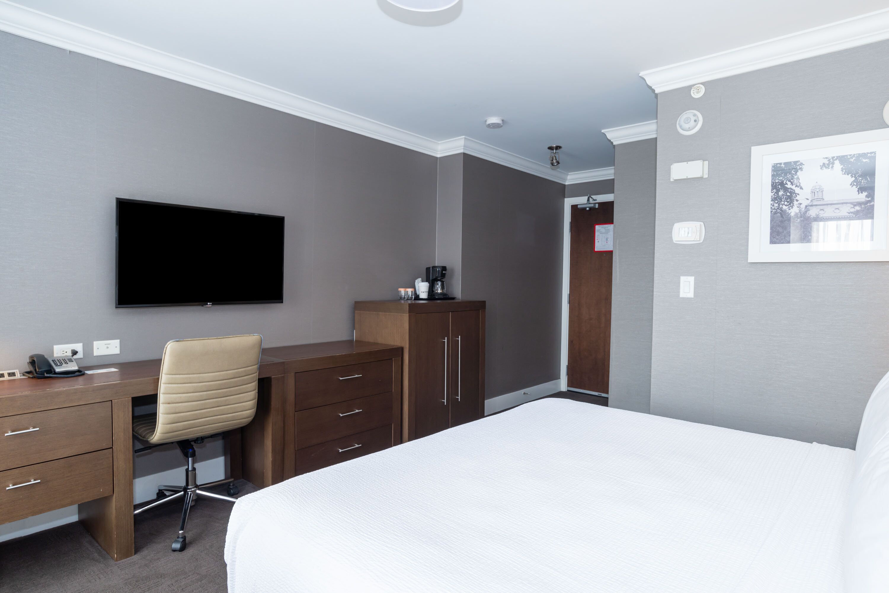 Quarto de hotel com uma cama, uma escrivaninha com cadeira, televisão de tela plana, guarda-roupa e um quadro na parede.