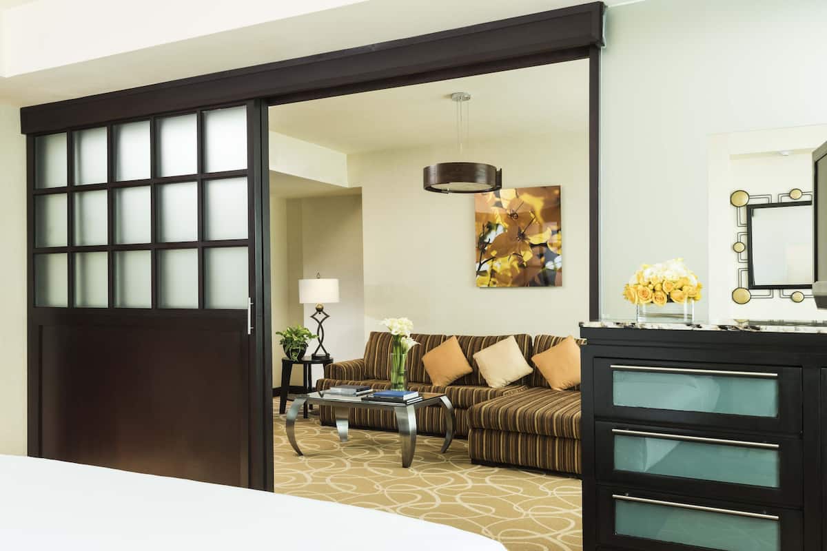 Quarto de hotel com uma cama, um sofá, uma mesa de centro, uma luminária, um vaso com flores e um espelho na parede.