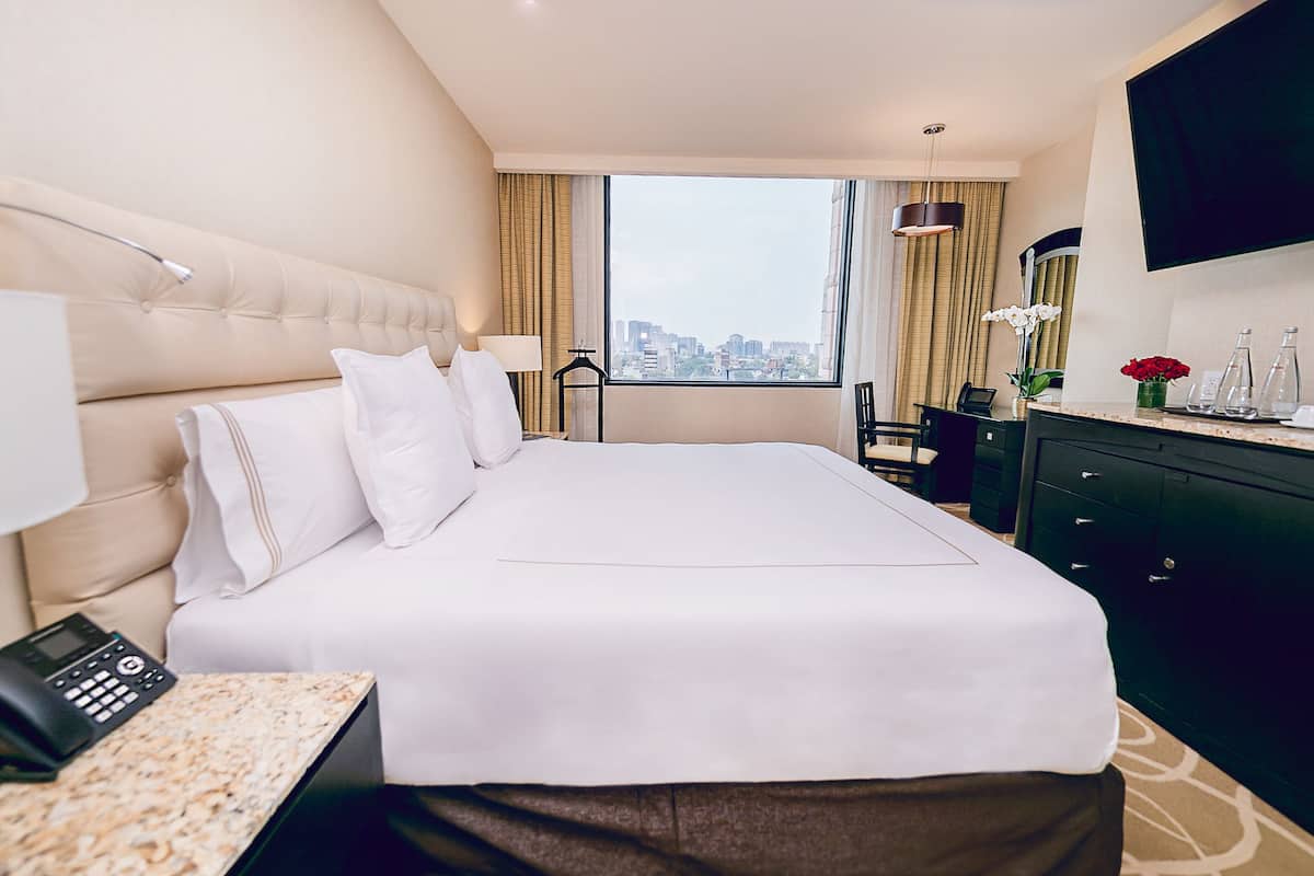 Quarto de hotel com uma cama grande, uma mesa com telefone, uma cadeira, uma TV e vista para a cidade.